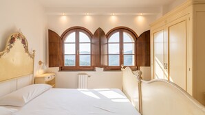 Quarto com cama, duas janelas com persianas de madeira, mesa de cabeceira com abajur e guarda-roupa.