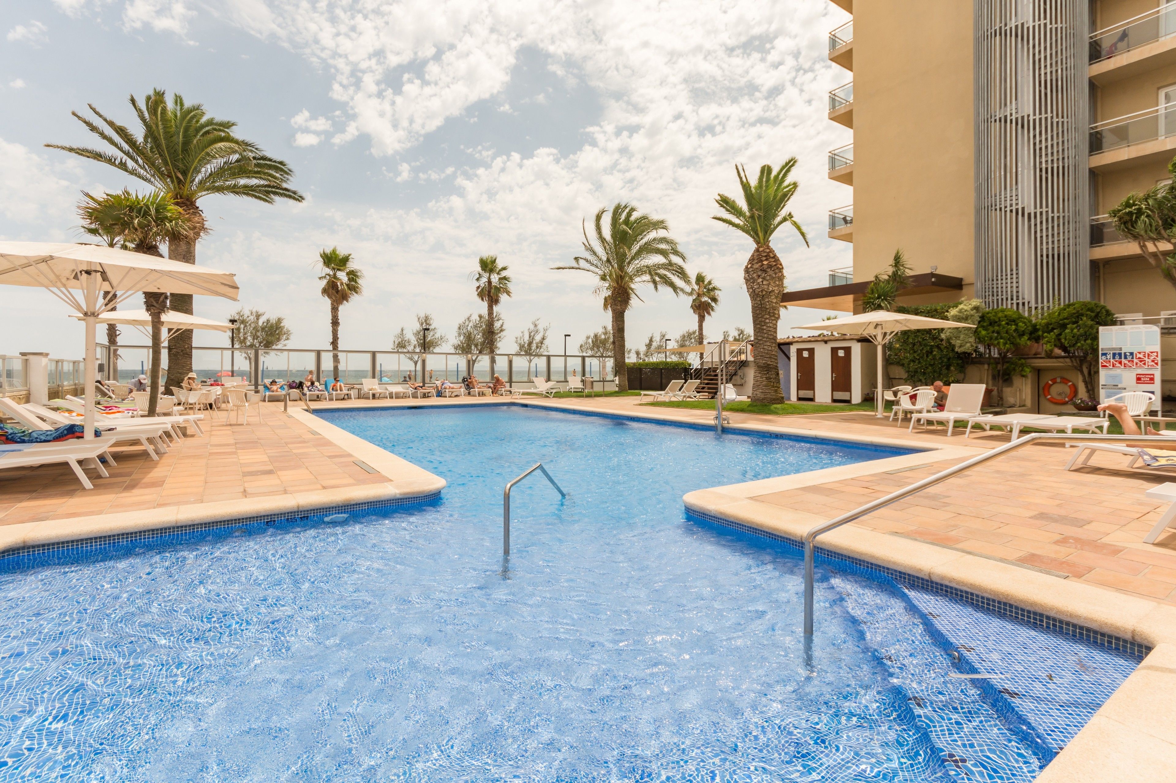 Piscina com espreguiçadeiras e palmeiras, cercada por um edifício com varandas.
