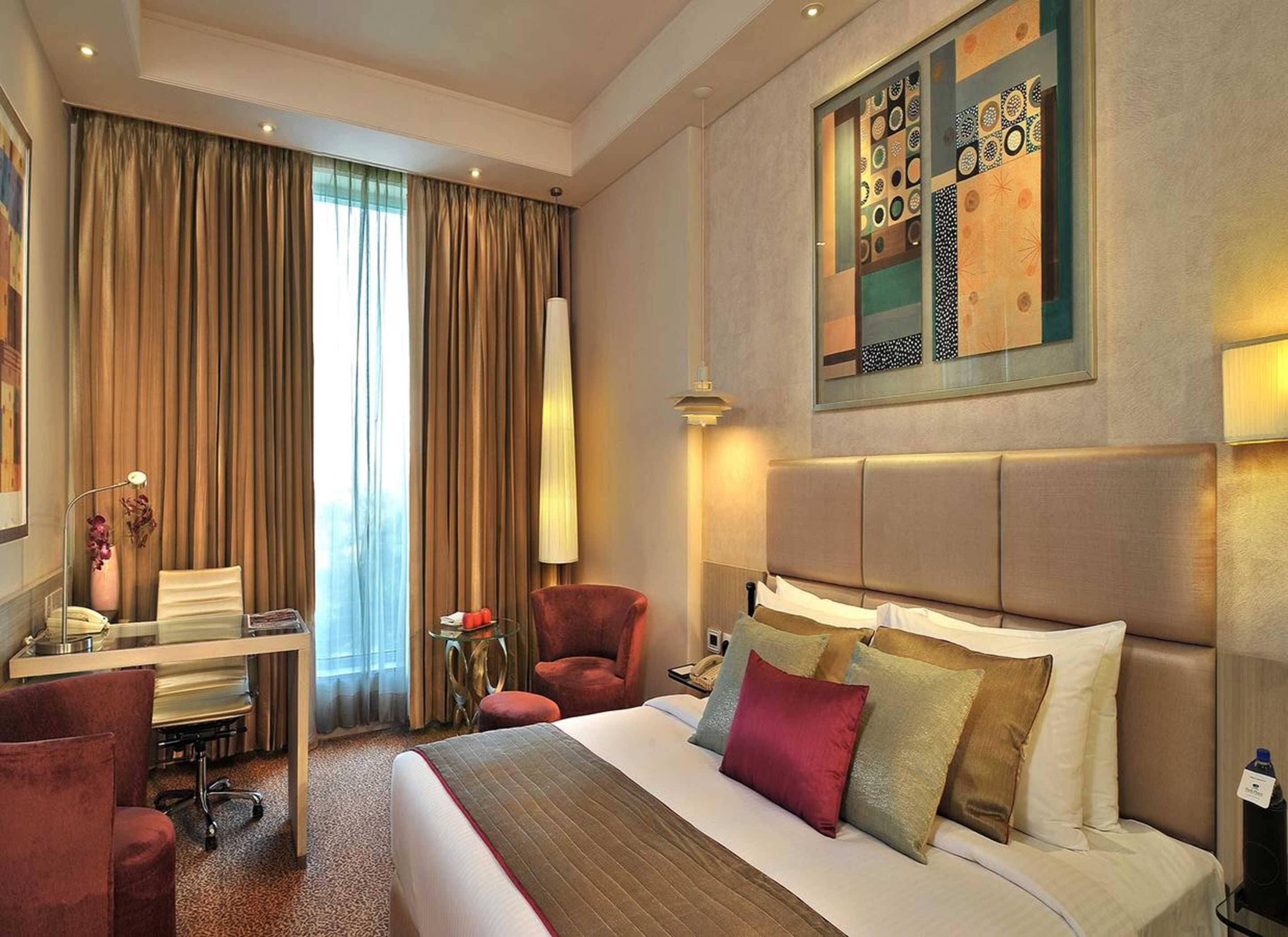 Quarto de hotel com cama grande, escrivaninha, cadeira e janela ampla com cortinas.