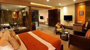 Quarto de hotel com uma cama grande, um espaço de estar com mesa de centro, televisão e banheiro com chuveiro e espelho.