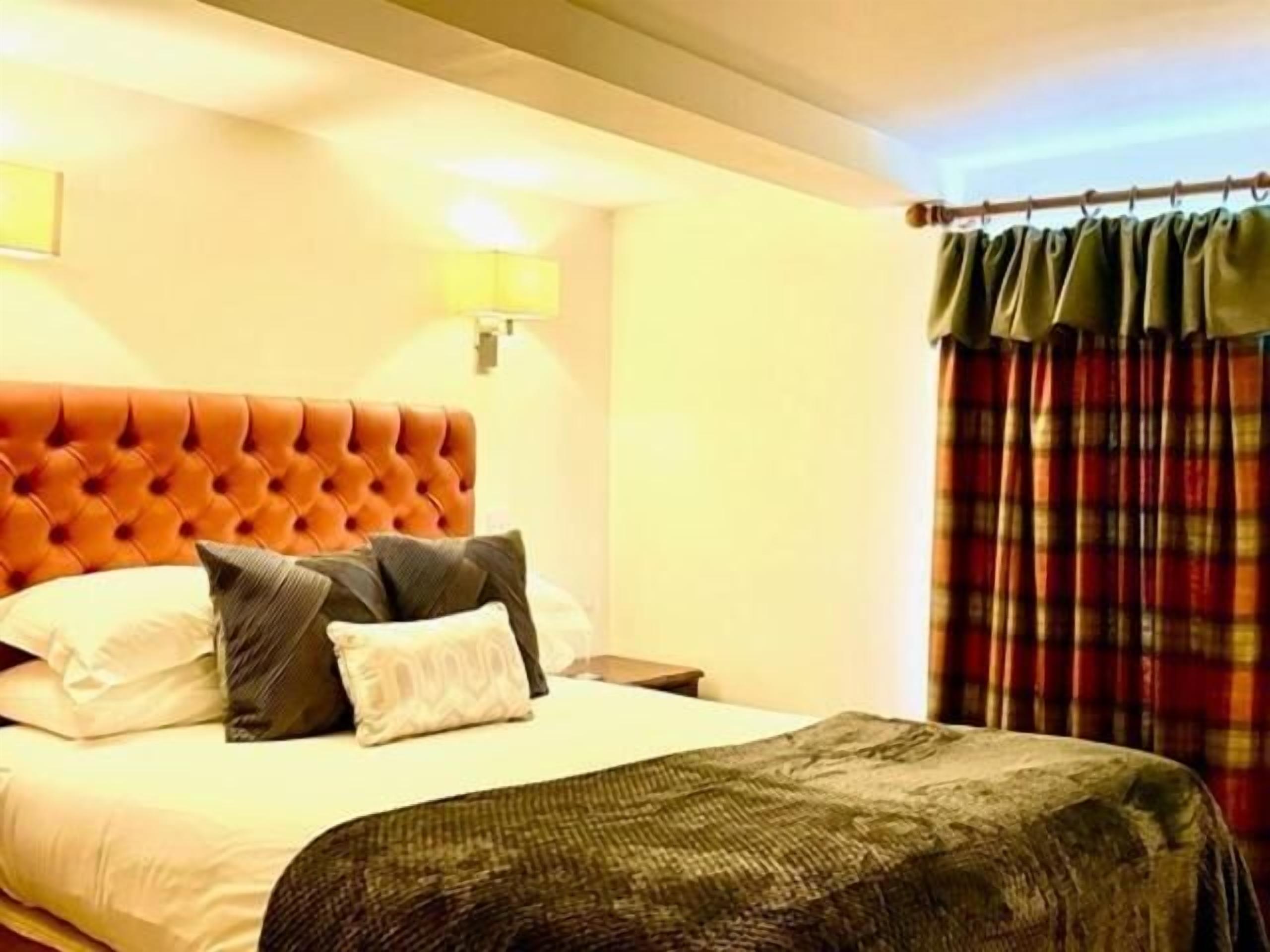Quarto de hotel com cabeceira estofada, cama com várias almofadas, luminária de cabeceira e cortina xadrez.