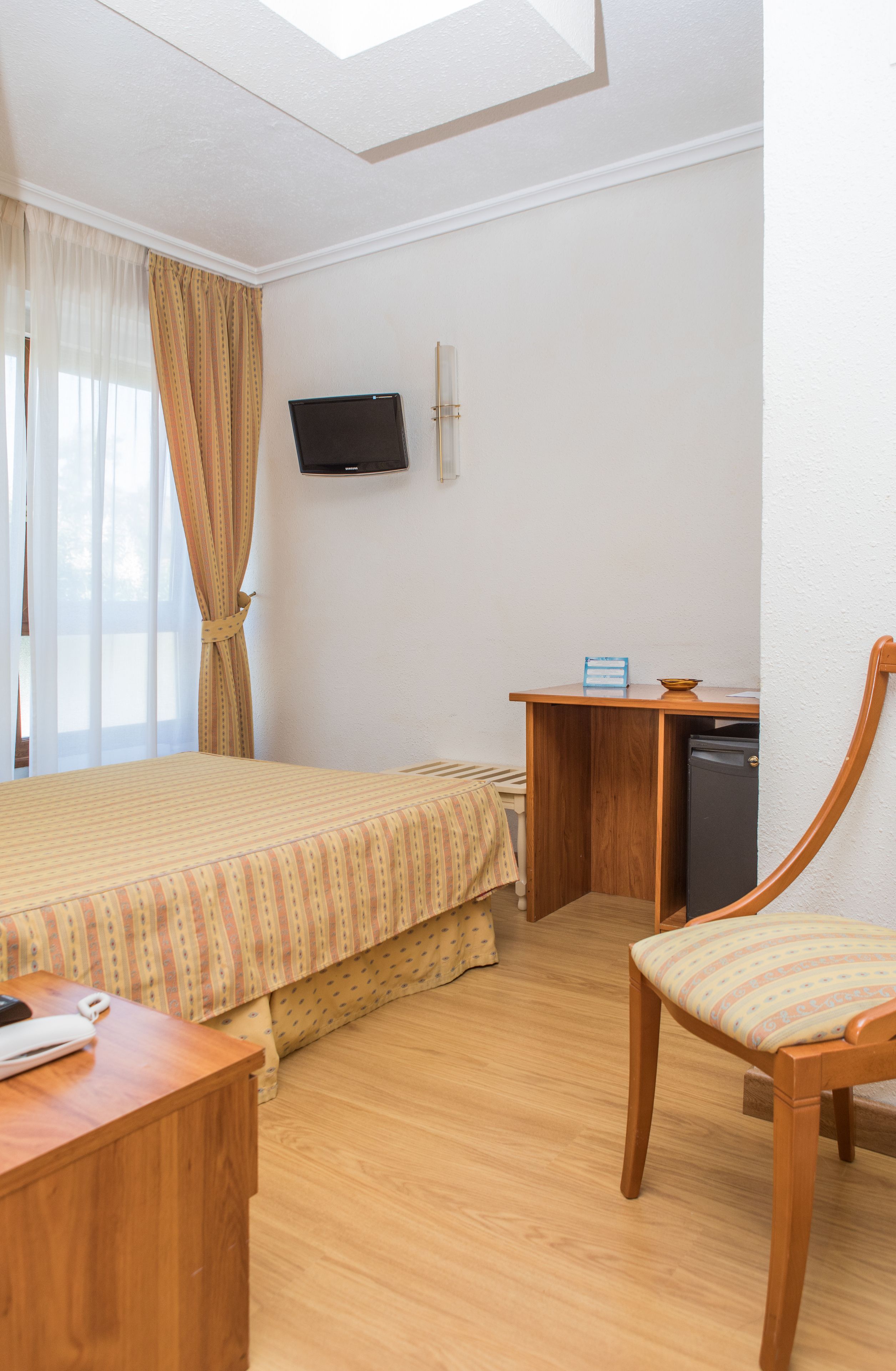 Quarto de hotel com cama, escrivaninha, cadeira, televisão e janela com cortinas.