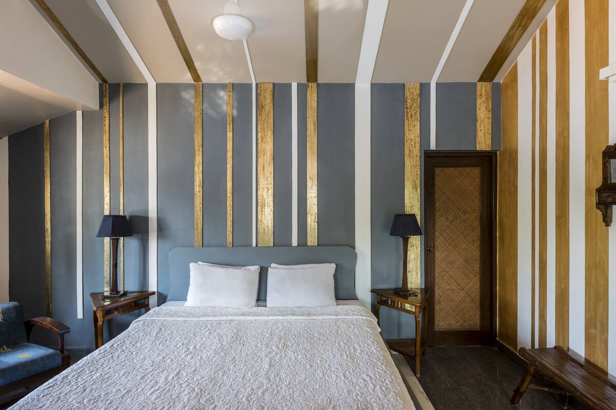 Quarto de hotel com uma cama grande, duas mesas de cabeceira, uma cadeira e uma porta que dá para outro cômodo.