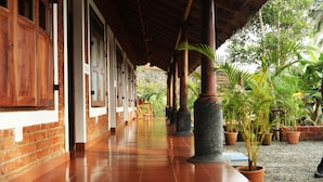 Um corredor coberto com paredes de tijolo e portas de madeira, que dá acesso a um jardim com plantas em vasos e uma área de estar.