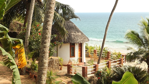 Um bangalô com telhado de palha e vista para o oceano, cercado por vegetação tropical e uma pequena área de varanda.