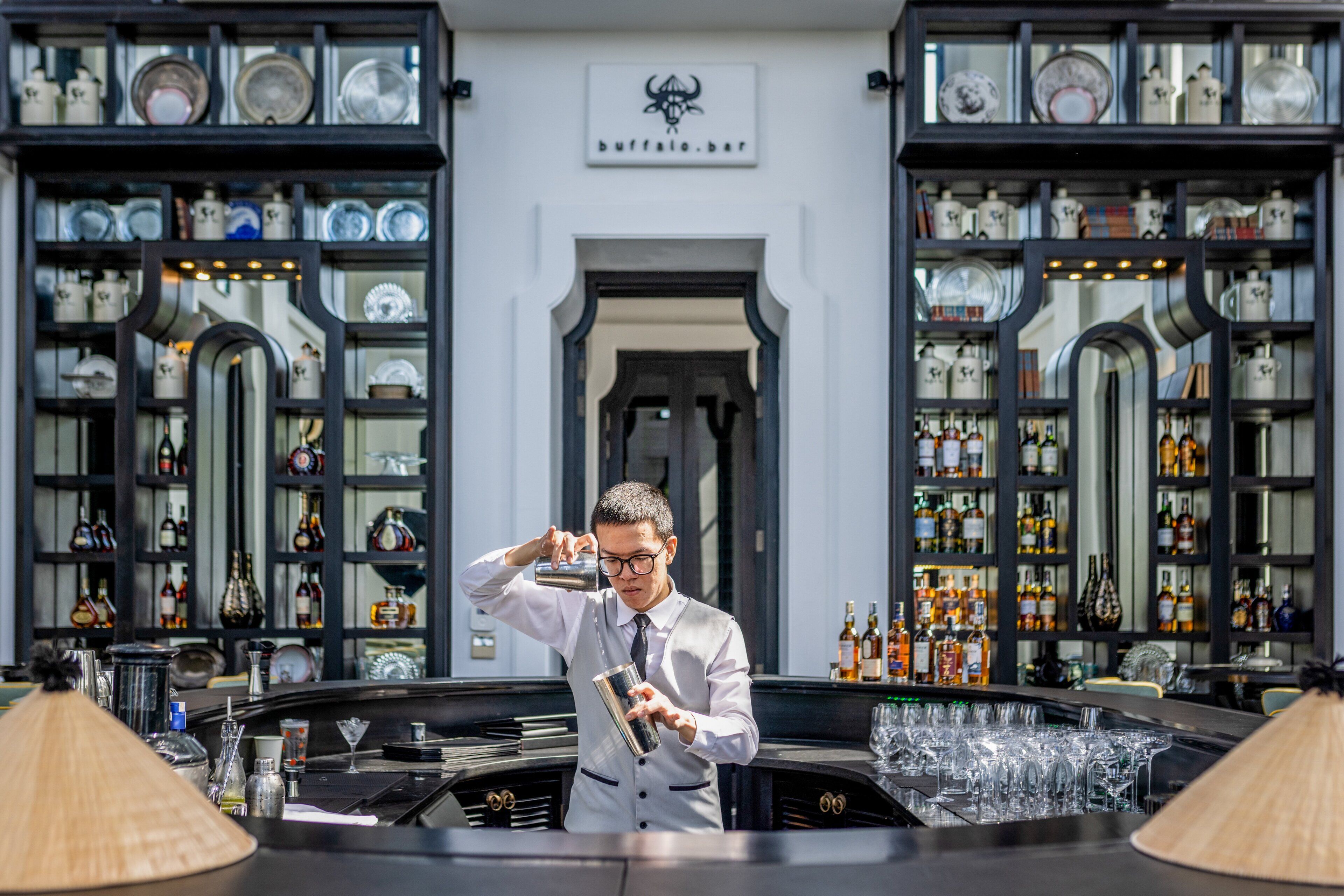 Um bartender em um bar, com prateleiras cheias de garrafas e copos atrás dele.