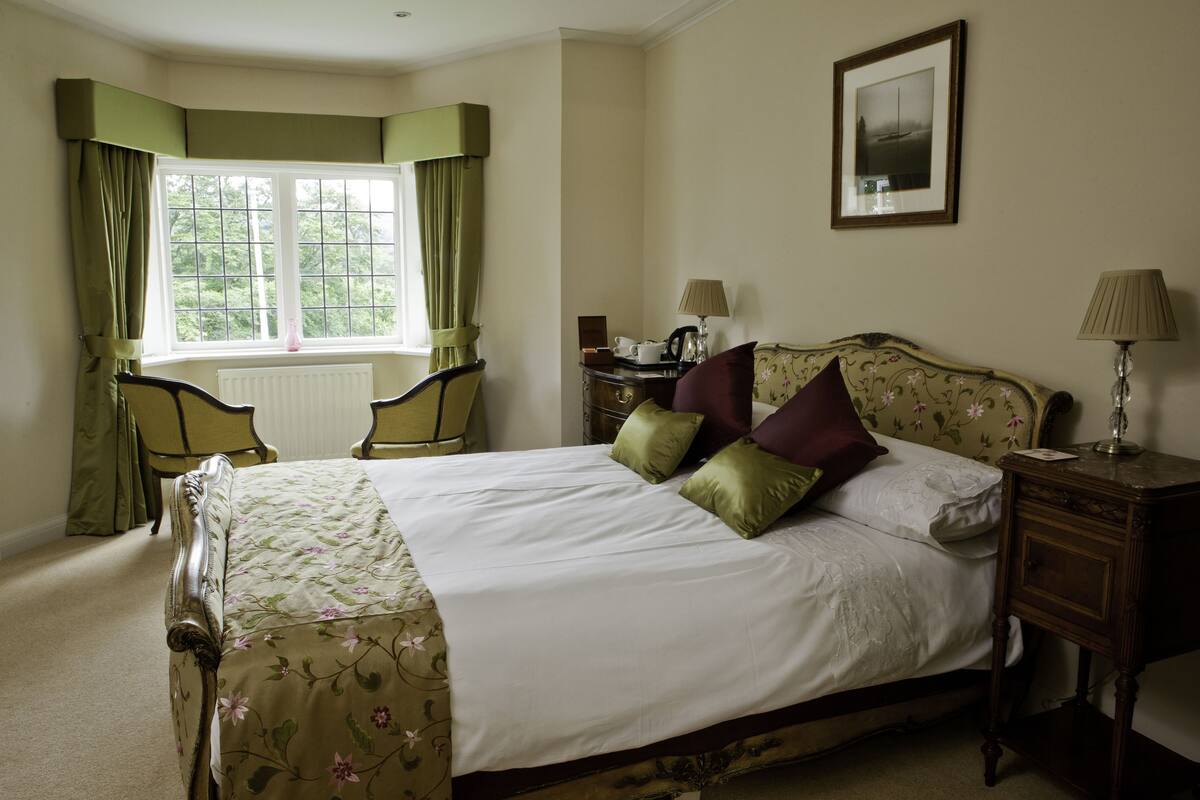 Quarto com cama grande, mesa de cabeceira, penteadeira, cadeira e janela com cortinas.