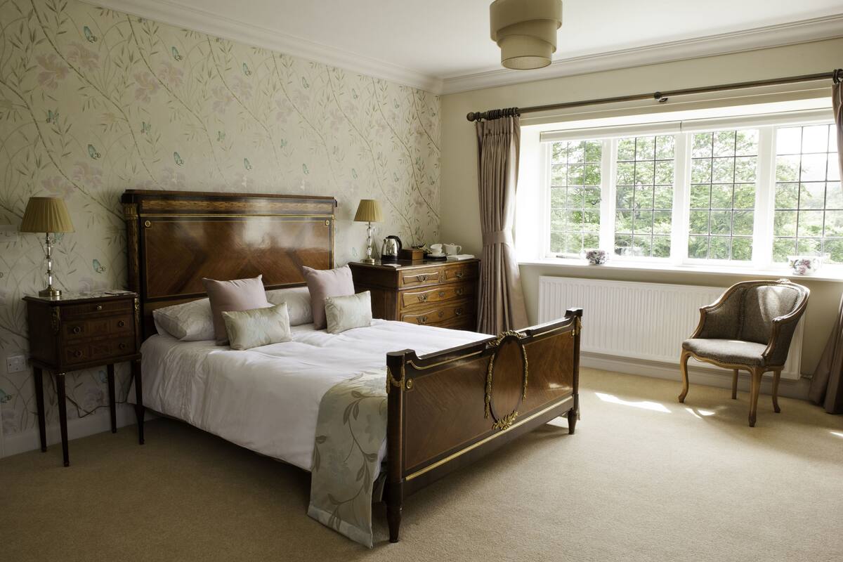 Quarto com cama grande, mesa de cabeceira, cômoda, cadeira e janela com cortinas.