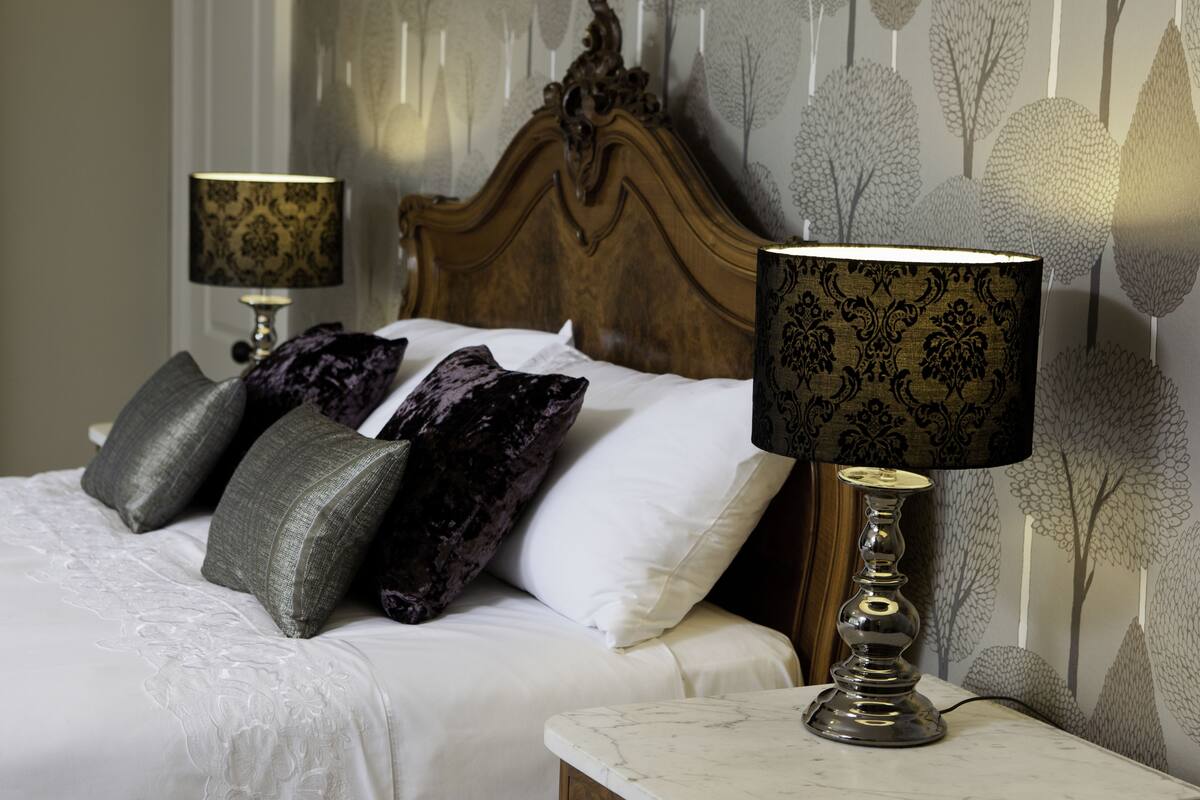 Cama bem arrumada com cabeceira de madeira, mesa de cabeceira de mármore e duas luminárias com cúpulas estampadas.