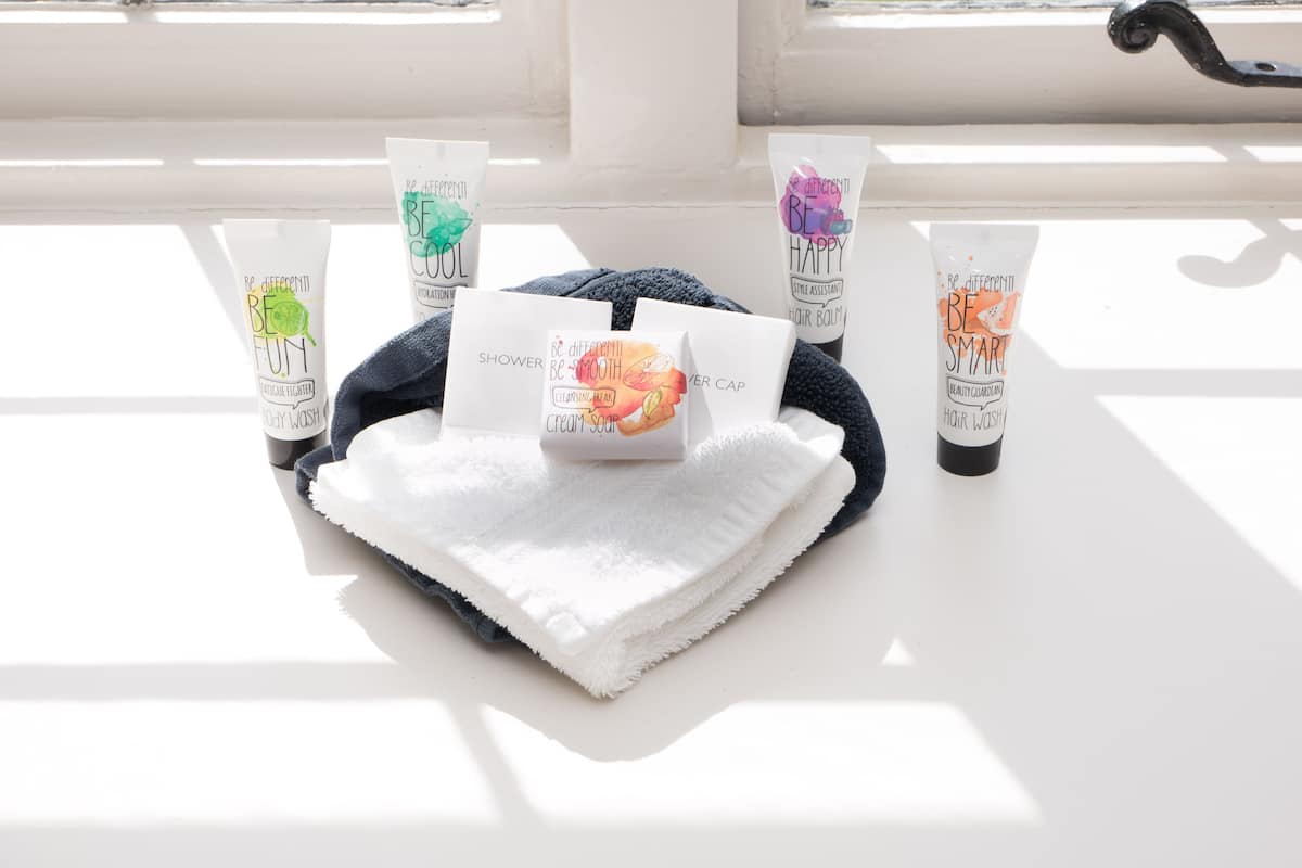 Uma bancada de banheiro com tampo branco, uma toalha azul e diversos produtos de higiene pessoal, incluindo banho cremoso, condicionador e touca de banho.
