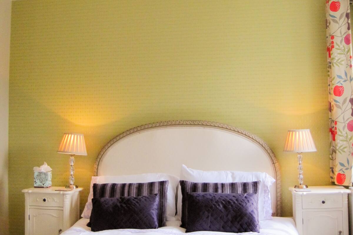 Cama bem arrumada com roupa de cama branca e roxa escura, duas luminárias de cabeceira e uma cortina com padrão floral.