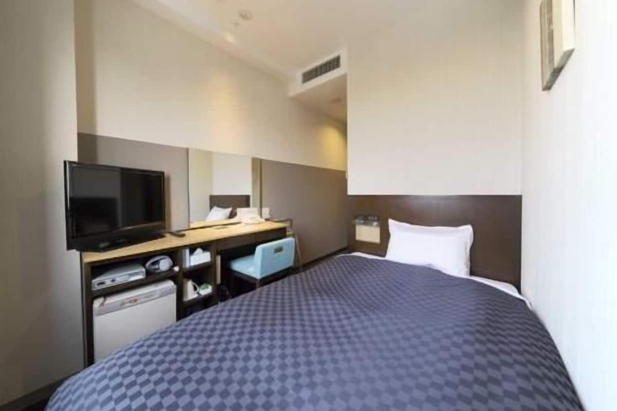 Quarto de hotel com uma cama, uma mesa de trabalho com televisão, uma kitchenette pequena e uma janela.
