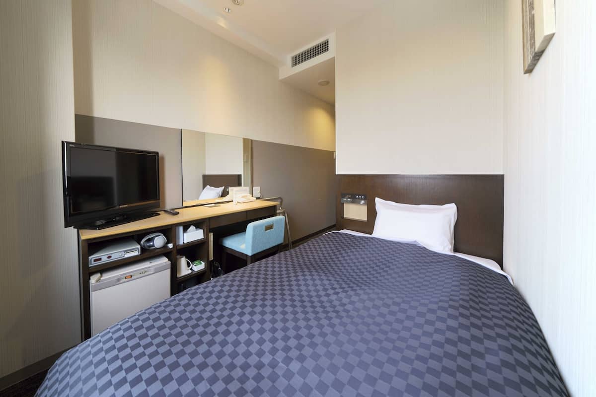 Quarto de hotel com cama, escrivaninha com televisão, espelho, pequena copa e janela.