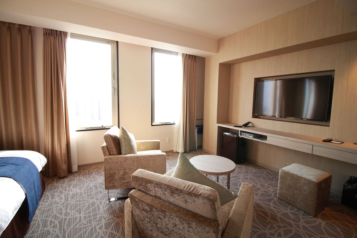 Quarto de hotel com uma cama, duas poltronas, uma mesinha, uma televisão e uma escrivaninha com um computador.