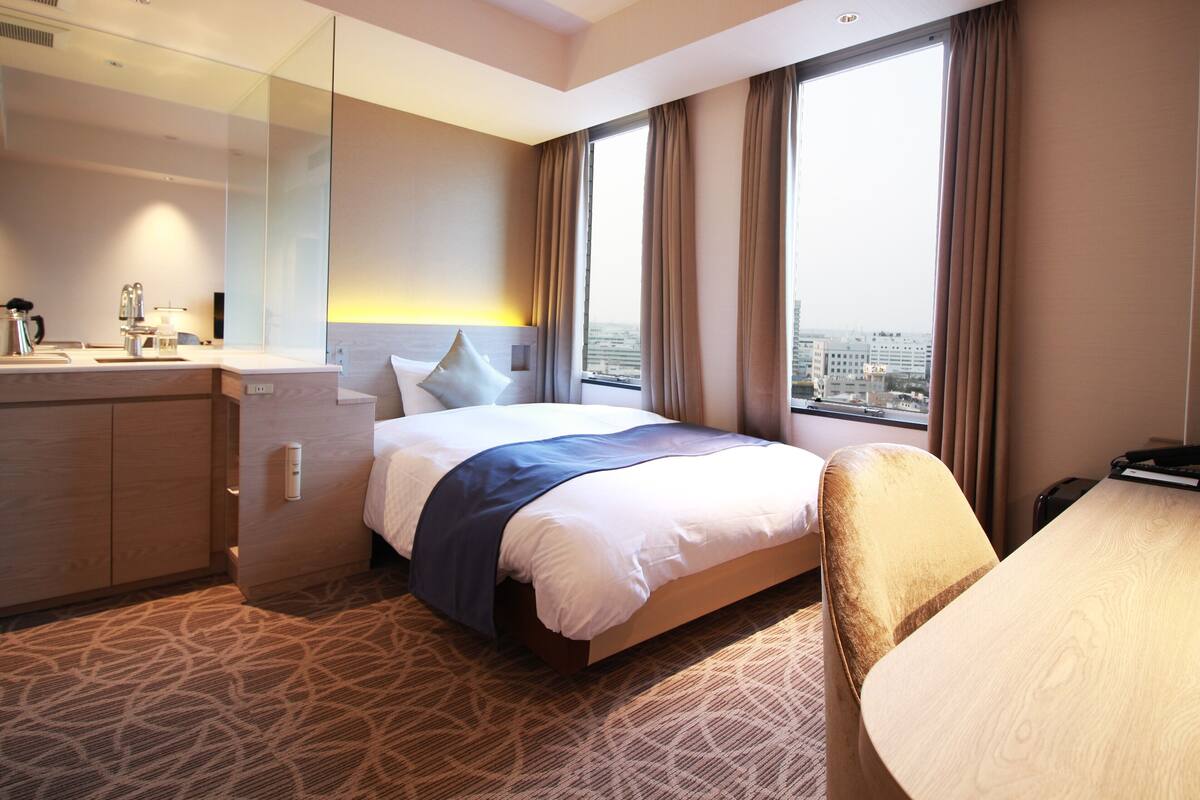 Quarto de hotel com cama, escrivaninha, espelho e vista da cidade.