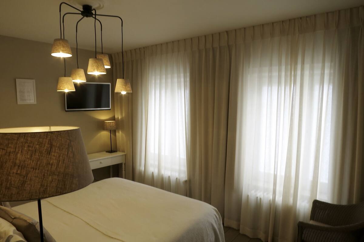 Quarto de hotel com uma cama, uma escrivaninha com luminária, uma televisão e uma cadeira.
