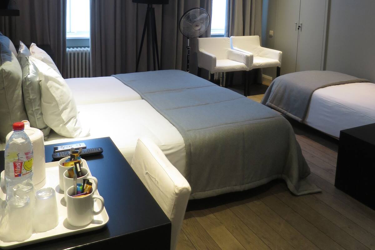 Quarto de hotel com duas camas, uma escrivaninha, uma cadeira, um ventilador, um abajur e uma janela com cortinas.