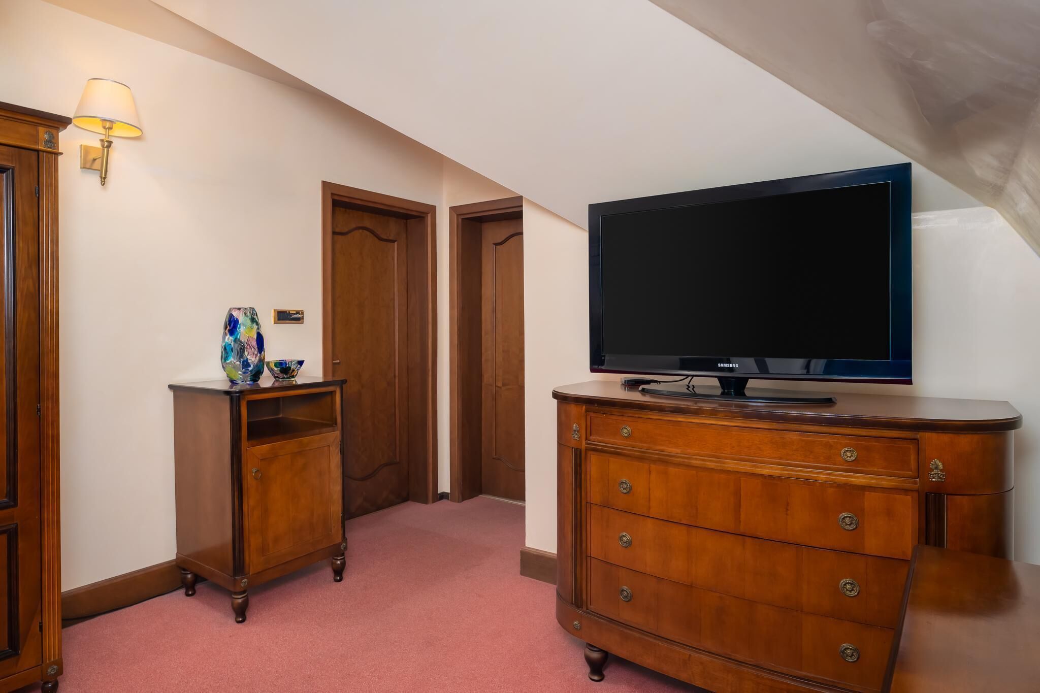 Quarto de hotel com cómoda de madeira, televisão em cima, mesinha com objetos decorativos e candeeiro fixado na parede.