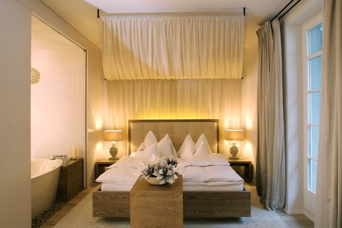 Uma cama de casal com cabeceira de madeira, duas mesas de cabeceira com abajures, uma janela grande com cortinas e uma banheira.