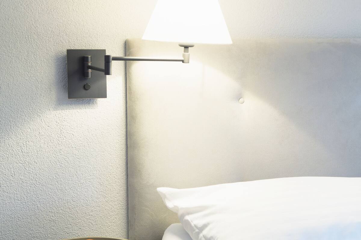 Uma cama bem arrumada com lençóis brancos, uma mesa de cabeceira com uma cesta de frutas e uma luminária na parede.
