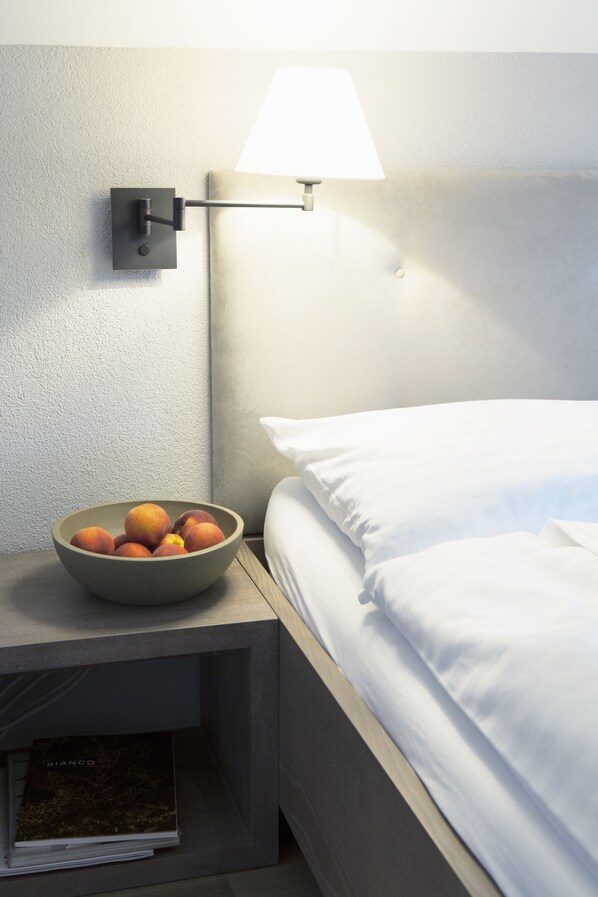 Uma cama bem arrumada com lençóis brancos, uma mesa de cabeceira com uma cesta de frutas e uma luminária na parede.