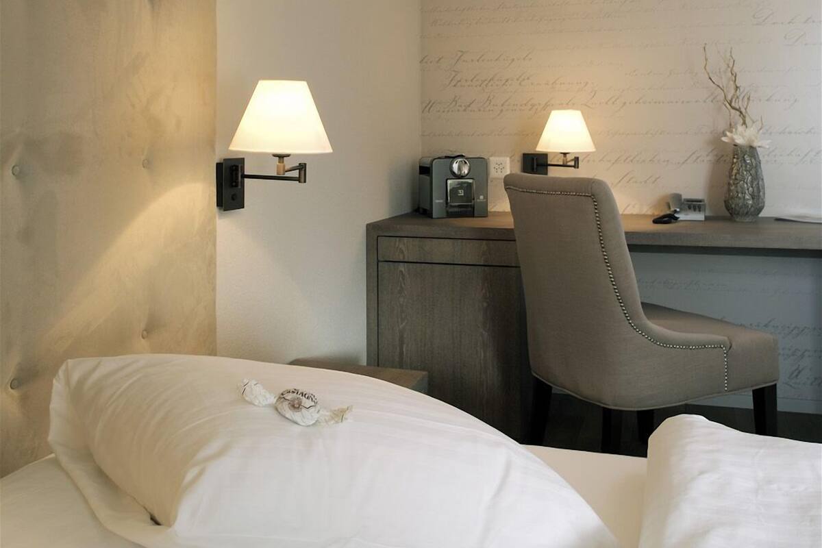 Quarto de hotel com uma cama, uma escrivaninha, duas luminárias e uma cadeira.