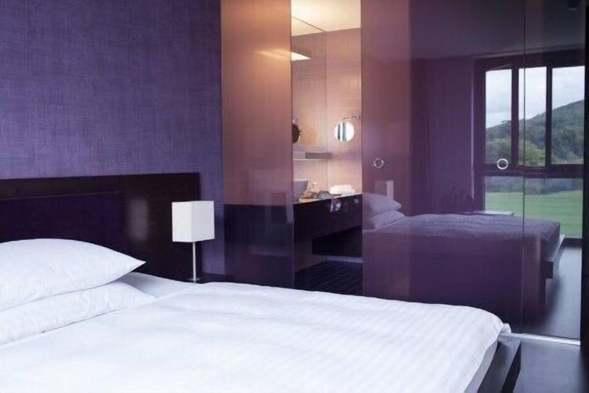 Quarto de hotel com uma cama grande, uma mesinha de cabeceira, um espelho e uma janela com vista para o exterior.