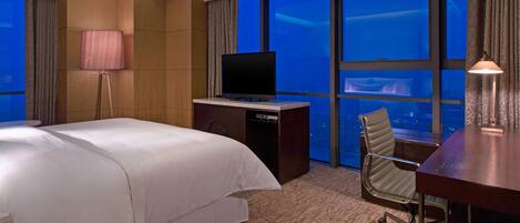 Quarto de hotel com cama, escrivaninha com televisão, cadeira e janelas amplas com vista para a cidade.