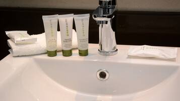 Uma pia de banheiro com três garrafas verdes de produtos Greenearth para banho e corpo, uma toalha branca e uma torneira.