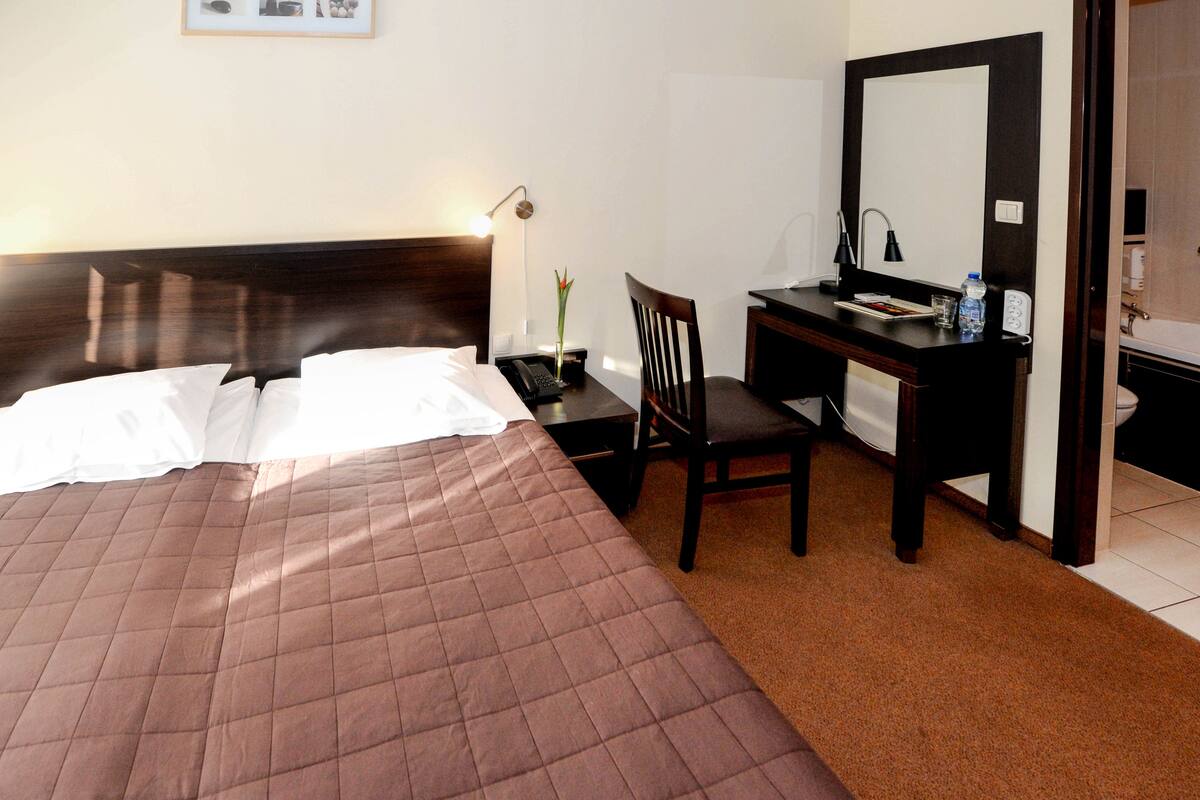 Quarto de hotel com cama, escrivaninha, cadeira, espelho e banheiro visível por uma porta aberta.