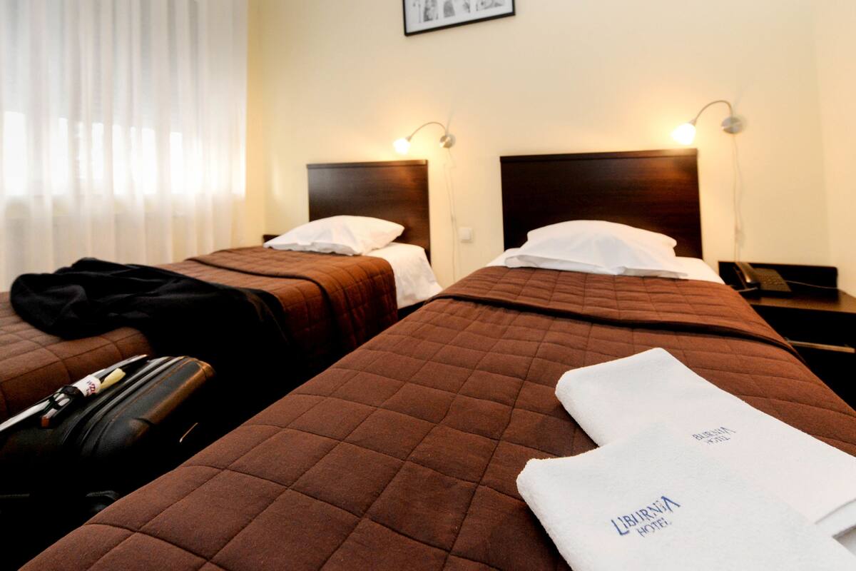Duas camas com colchas marrons, uma mala preta e uma toalha branca com a inscrição "LIBIRNE HOTEL".