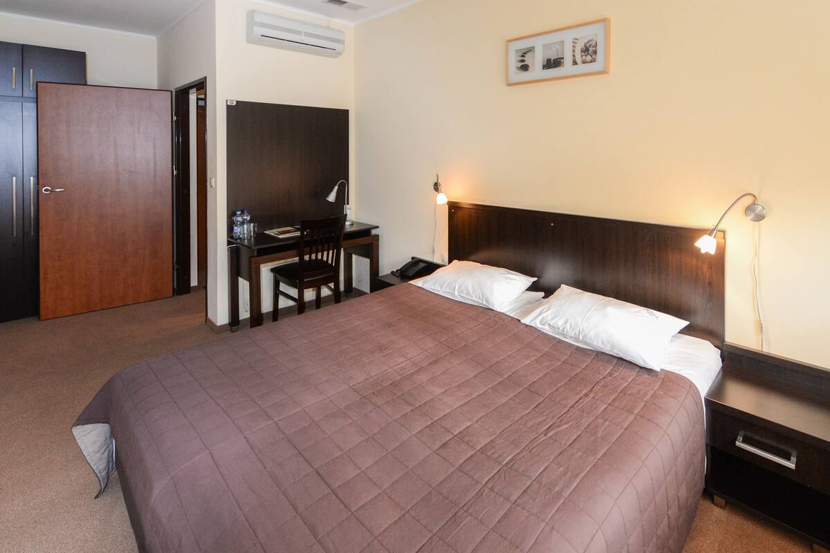 Quarto de hotel com uma cama grande, uma escrivaninha com cadeira, uma mesinha de cabeceira e um abajur fixado na parede.