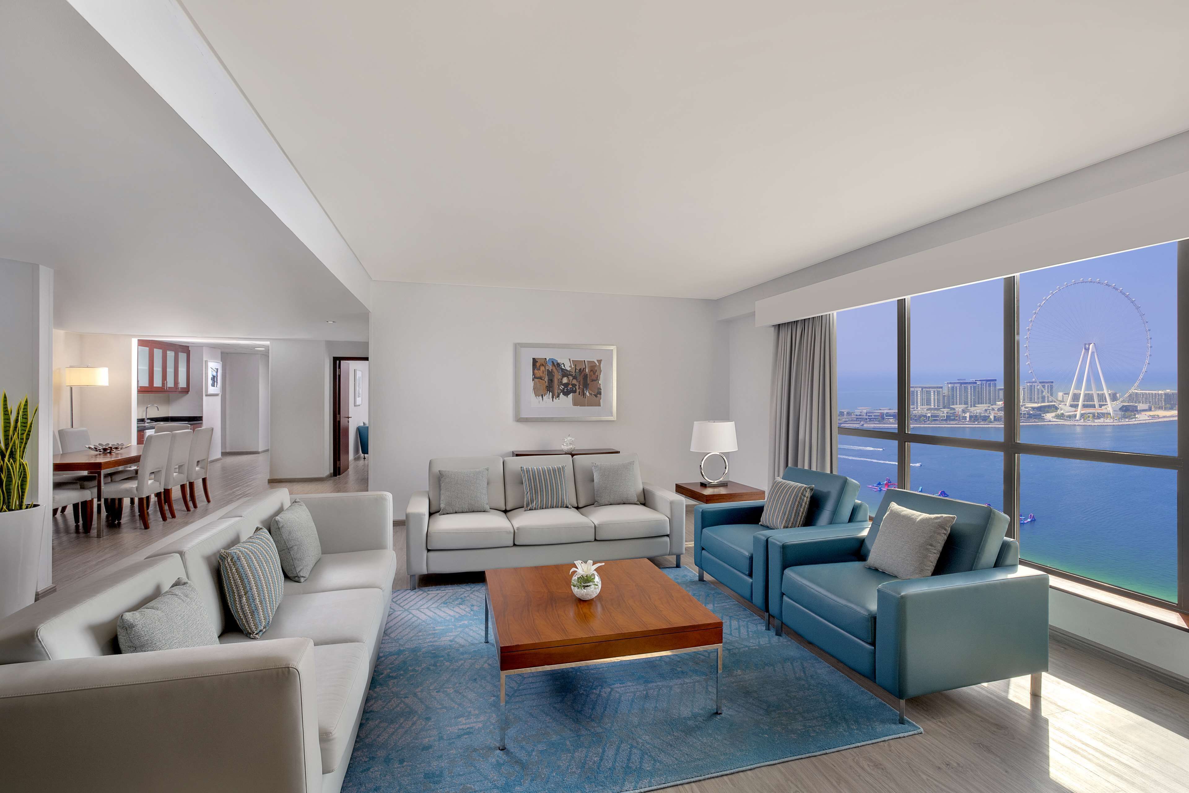 Quarto de hotel moderno com uma grande janela que oferece vista para uma roda gigante e para a água. Há um sofá, uma poltrona azul e uma mesa de centro de madeira.