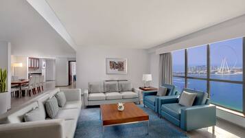 Quarto de hotel moderno com uma grande janela que oferece vista para uma roda gigante e para a água. Há um sofá, uma poltrona azul e uma mesa de centro de madeira.
