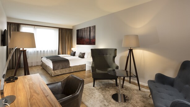 Quarto de hotel com cama, escrivaninha, cadeira, luminária e um quadro na parede.