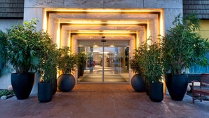 Entrada de um edifício moderno com porta de vidro, cercada por grandes vasos de plantas e iluminada por luzes no teto.