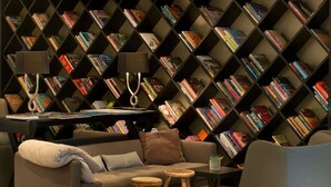 Uma sala de estar moderna com um sofá cinzento, dois taburetes de madeira, uma mesa pequena e uma estante repleta de livros.