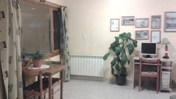 Uma sala de estar com uma mesa de madeira, um vaso de planta, uma escrivaninha com monitor e quadros na parede.