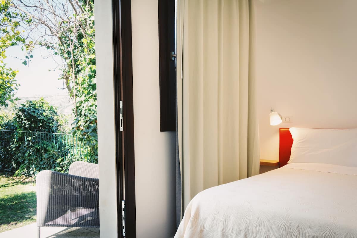 Quarto de hotel com cama, mesa de cabeceira com abajur, cadeira e vista para o verde lá fora.