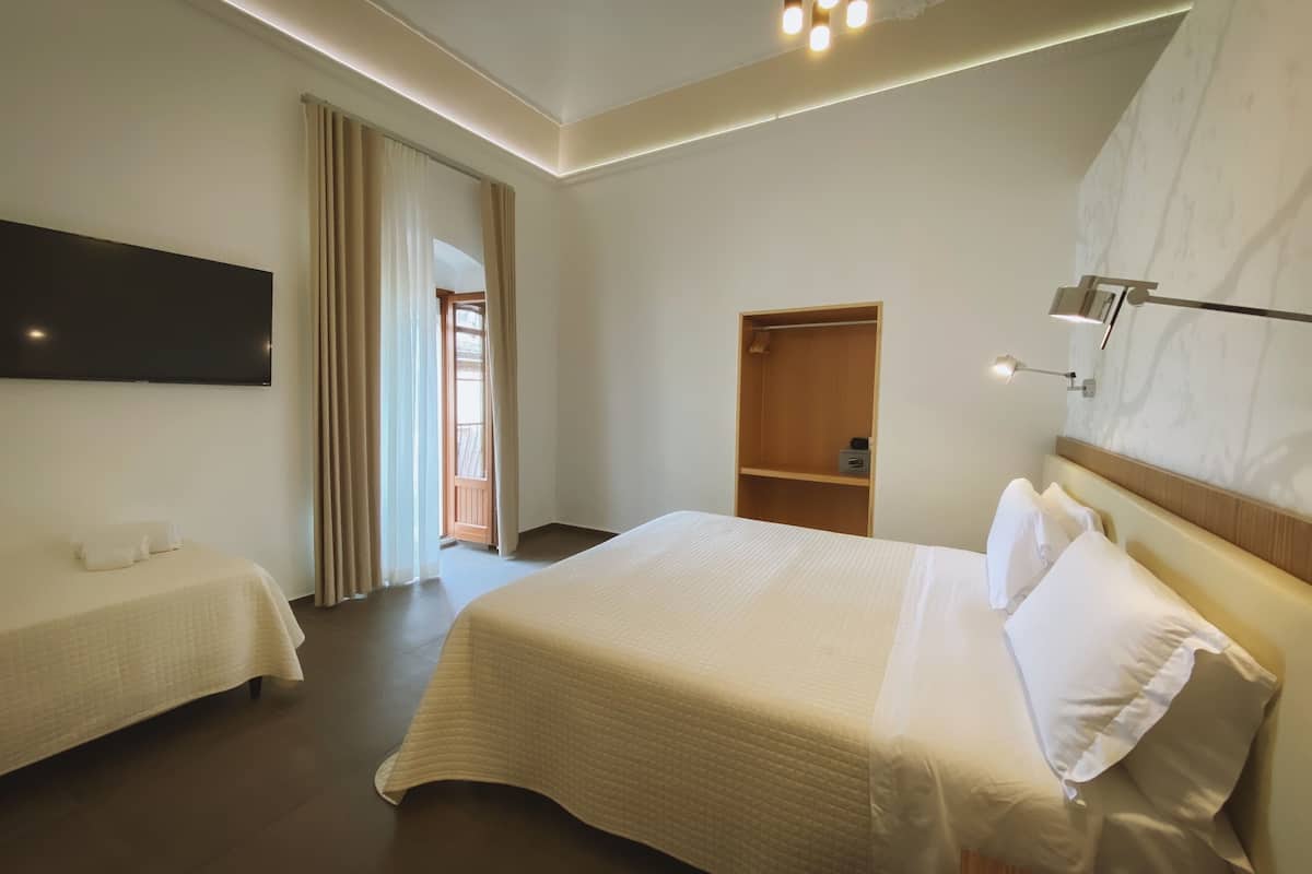 Quarto de hotel com uma cama grande, uma cama menor, uma TV fixada na parede, um guarda-roupa e uma porta que dá para uma varanda.
