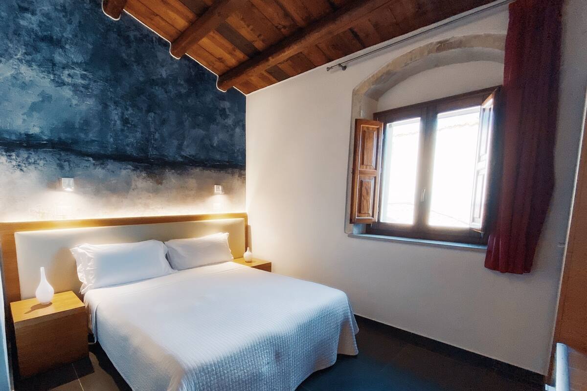 Quarto com cama, mesa de cabeceira e janela com cortinas.