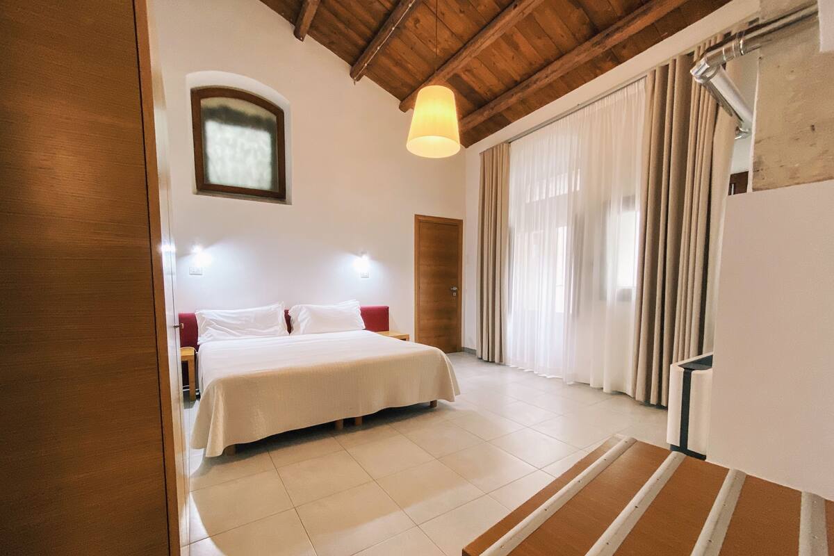Quarto com cama, teto de madeira, janela com cortinas e luminária.
