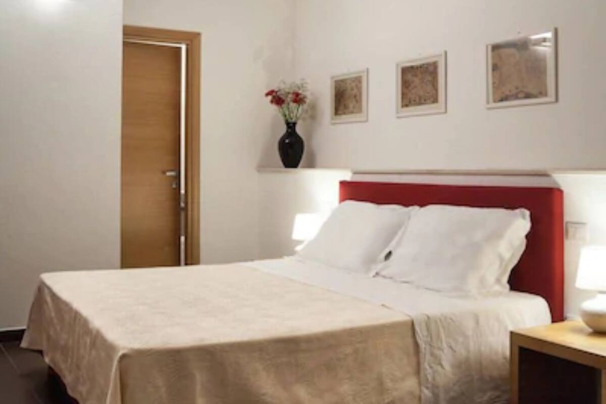 Cama bem arrumada com cabeceira vermelha, mesa de cabeceira com abajur e vaso de flores na parede.