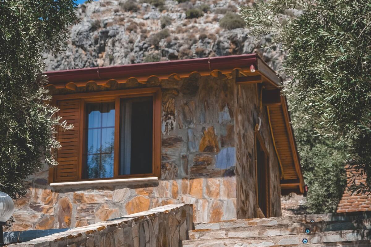 Uma escadaria de pedra leva a uma cabana de madeira com telhado vermelho, rodeada por oliveiras e terreno rochoso.