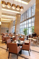 Ampla sala de jantar de hotel com várias mesas e cadeiras, grandes janelas e luminárias modernas.