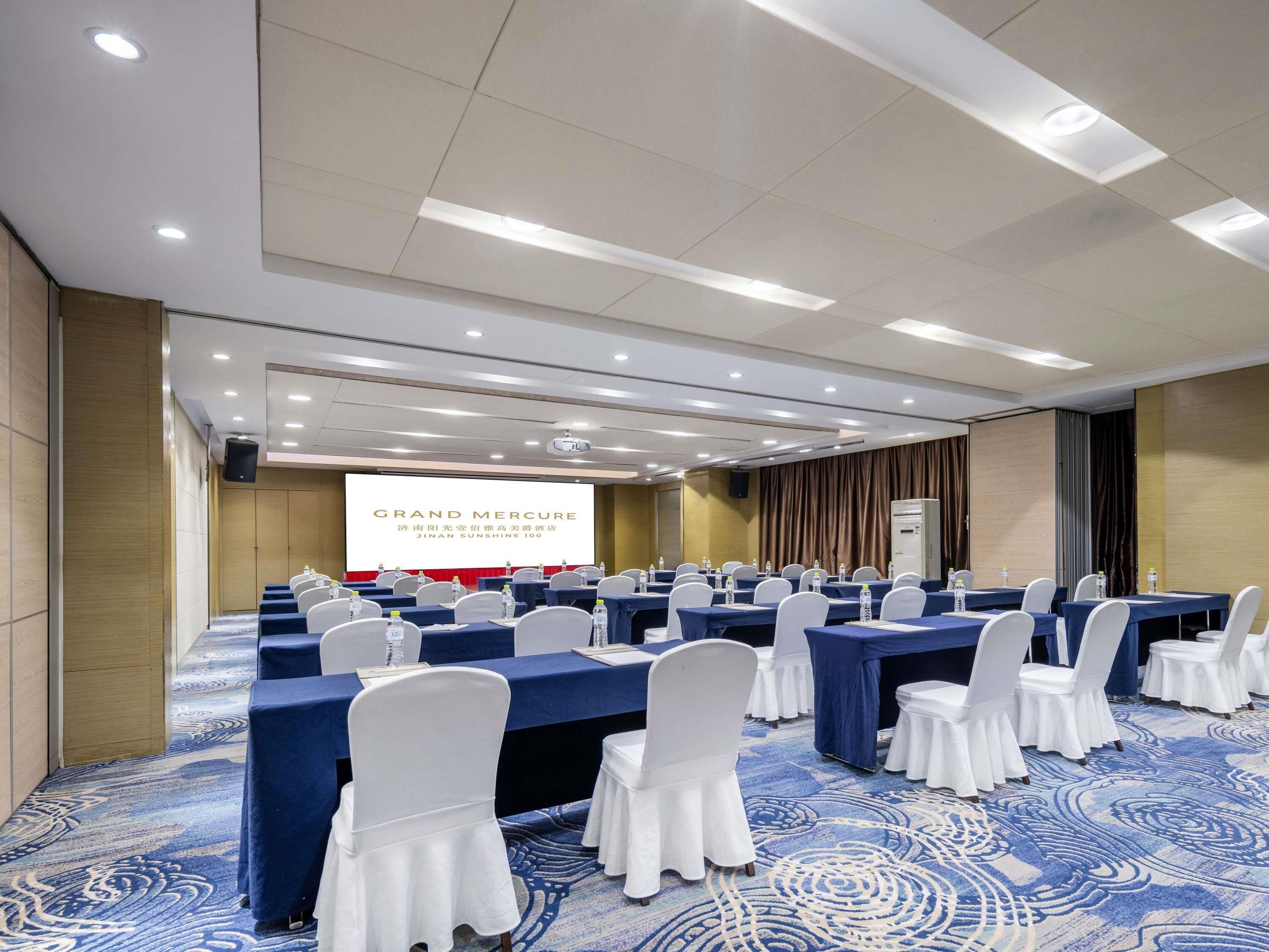 Uma sala de conferências com fileiras de mesas, cadeiras brancas e uma grande tela exibindo 'GRAND MERCURE'.
