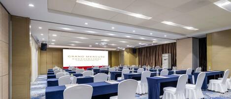 Uma sala de conferências com fileiras de mesas, cadeiras brancas e uma grande tela exibindo 'GRAND MERCURE'.
