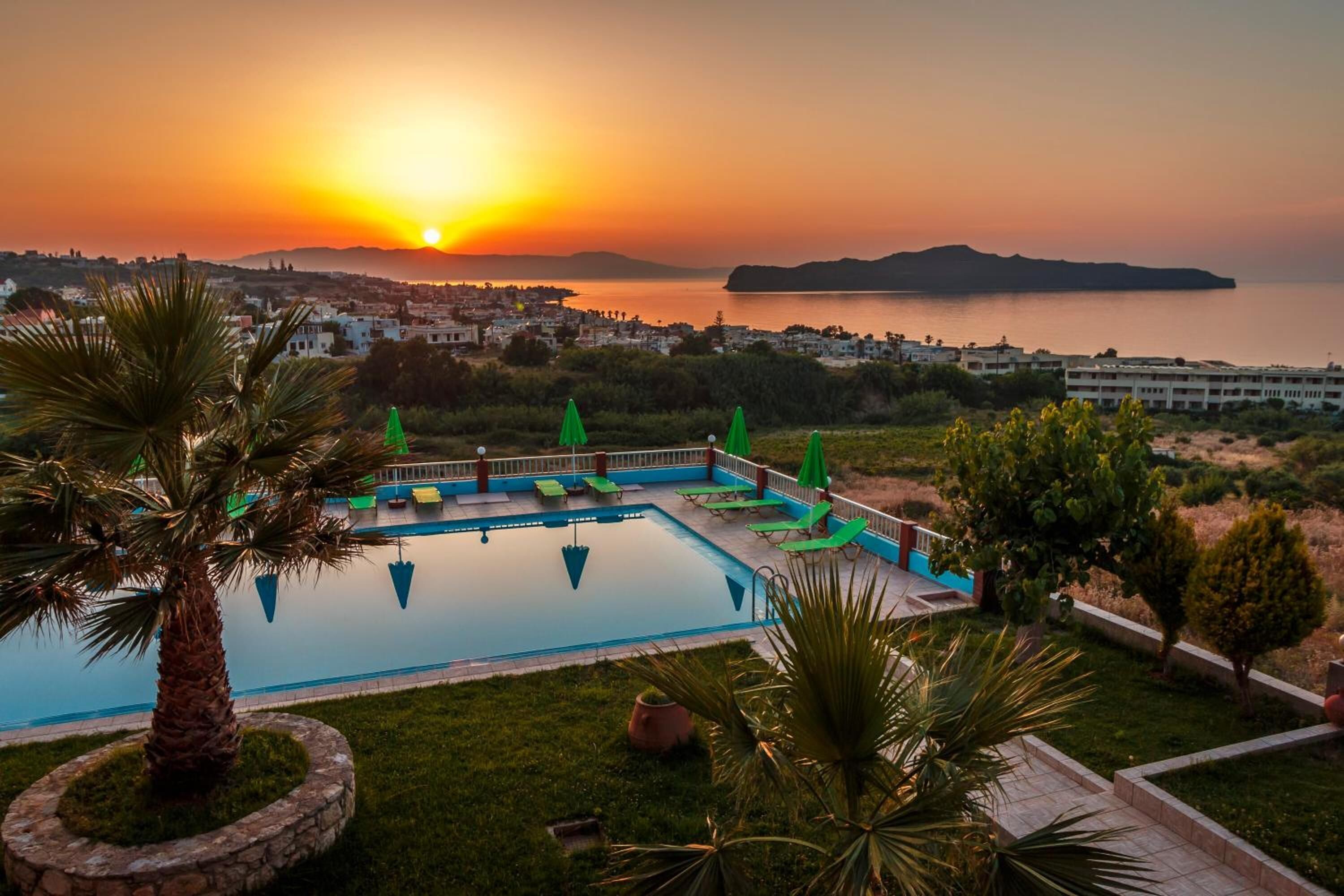 Piscina com espreguiçadeiras e guarda-sóis, cercada por vegetação e com vista para a paisagem urbana e um corpo d'água ao pôr do sol.