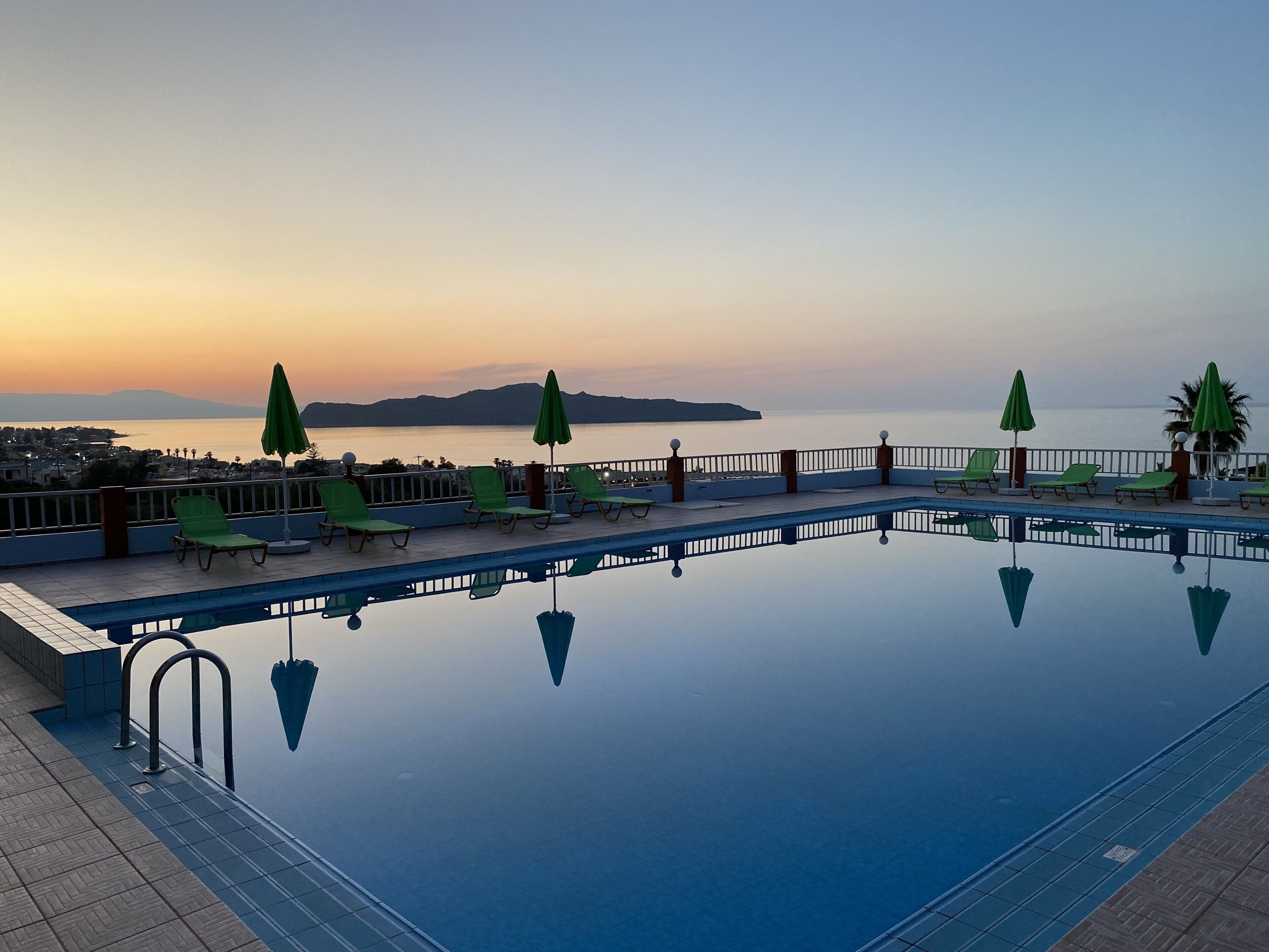 Piscina com espreguiçadeiras e guarda-sóis verdes, rodeada por um deck ladrilhado, com vista para um corpo d'água calmo e colinas ao longe ao pôr do sol.