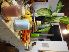 Uma mesa de jantar com frutas variadas, um jarro e um vaso com planta.
